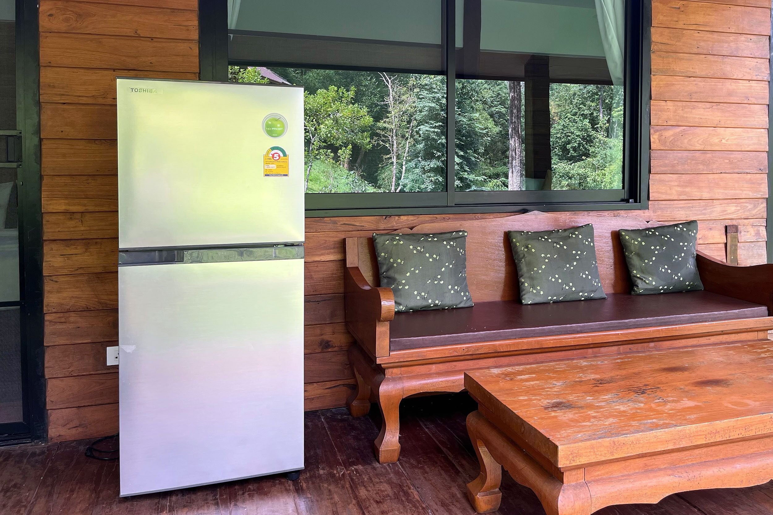Interior de cabana de madeira com frigorífico branco, banco de madeira com almofadas verdes e uma grande janela com vista para árvores.