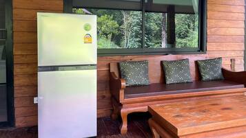 Interior de cabana de madeira com frigorífico branco, banco de madeira com almofadas verdes e uma grande janela com vista para árvores.