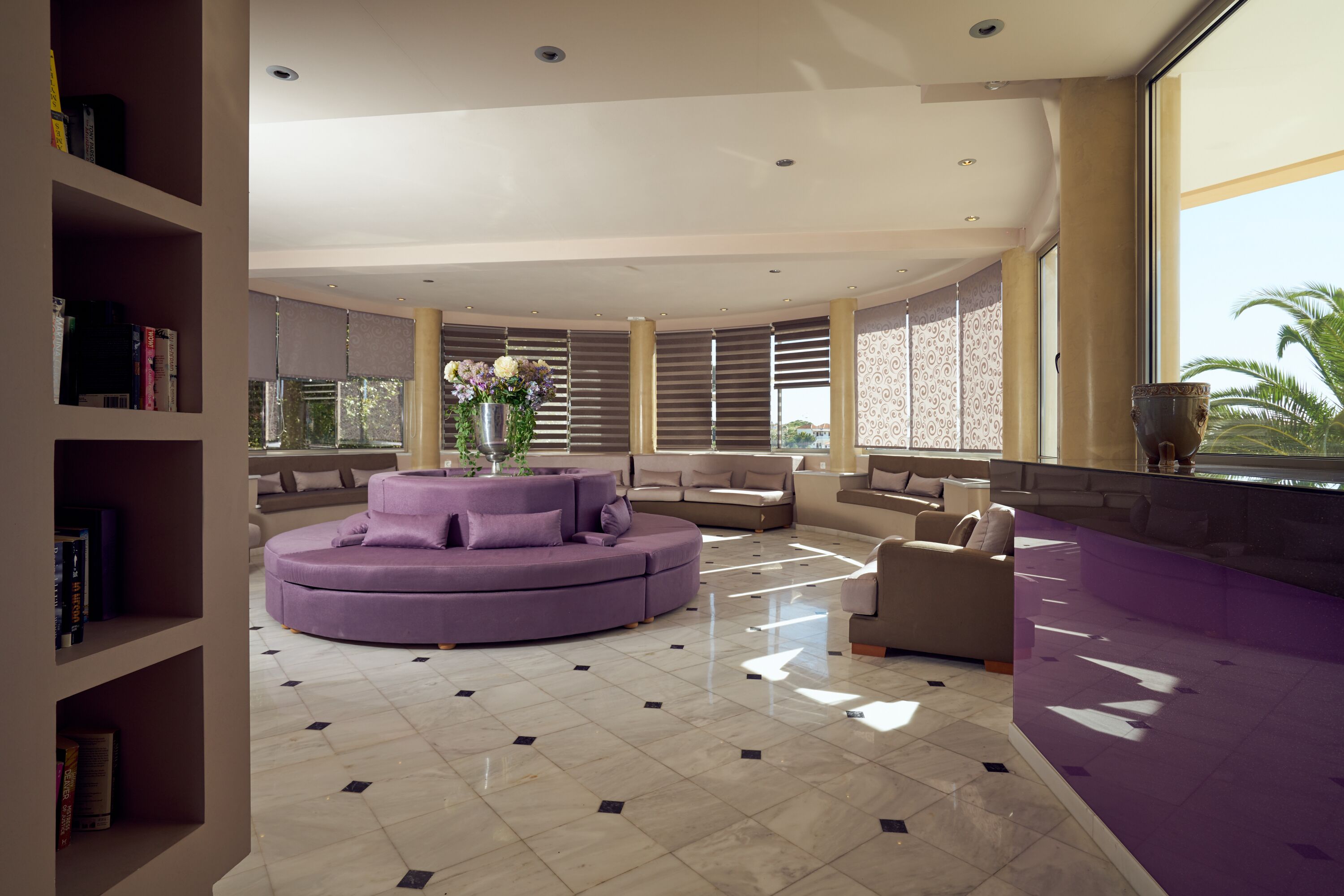 Uma sala moderna com um amplo sofá em formato de L, um sofá roxo redondo e uma poltrona bege. Há uma estante à esquerda e uma janela grande à direita.