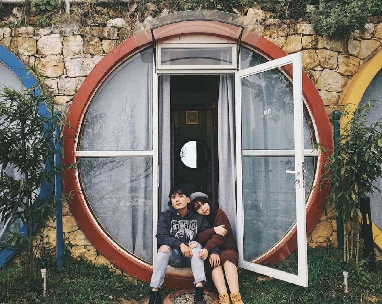 Janela circular com parede de pedra ao fundo, uma pessoa sentada no interior e uma porta com janela circular.