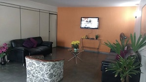 Uma sala de estar moderna com um sofá, uma poltrona estampada, uma mesa redonda pequena e uma televisão presa a uma parede laranja.