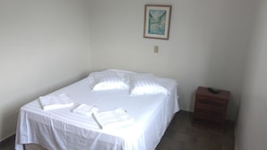 Quarto simples e limpo com uma cama de solteiro, uma pequena mesa de cabeceira de madeira e um quadro na parede.