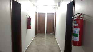 Um corredor com dois extintores fixados nas paredes, uma porta de madeira no fim e chão de azulejos.