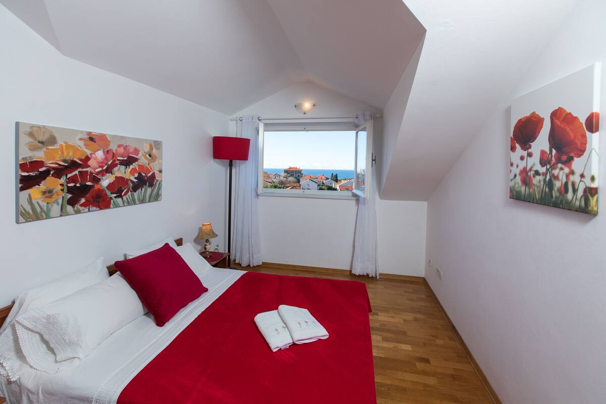 Quarto com uma cama grande, um cobertor vermelho, almofadas brancas e duas pinturas de flores nas paredes.