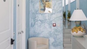 Um corredor com parede de mármore azul, um quadro emoldurado, uma poltrona pequena e um abajur sobre uma mesa.