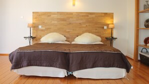 Uma cama de casal com cabeceira de madeira, duas luminárias de apoio e um pequeno aparador com objetos decorativos.