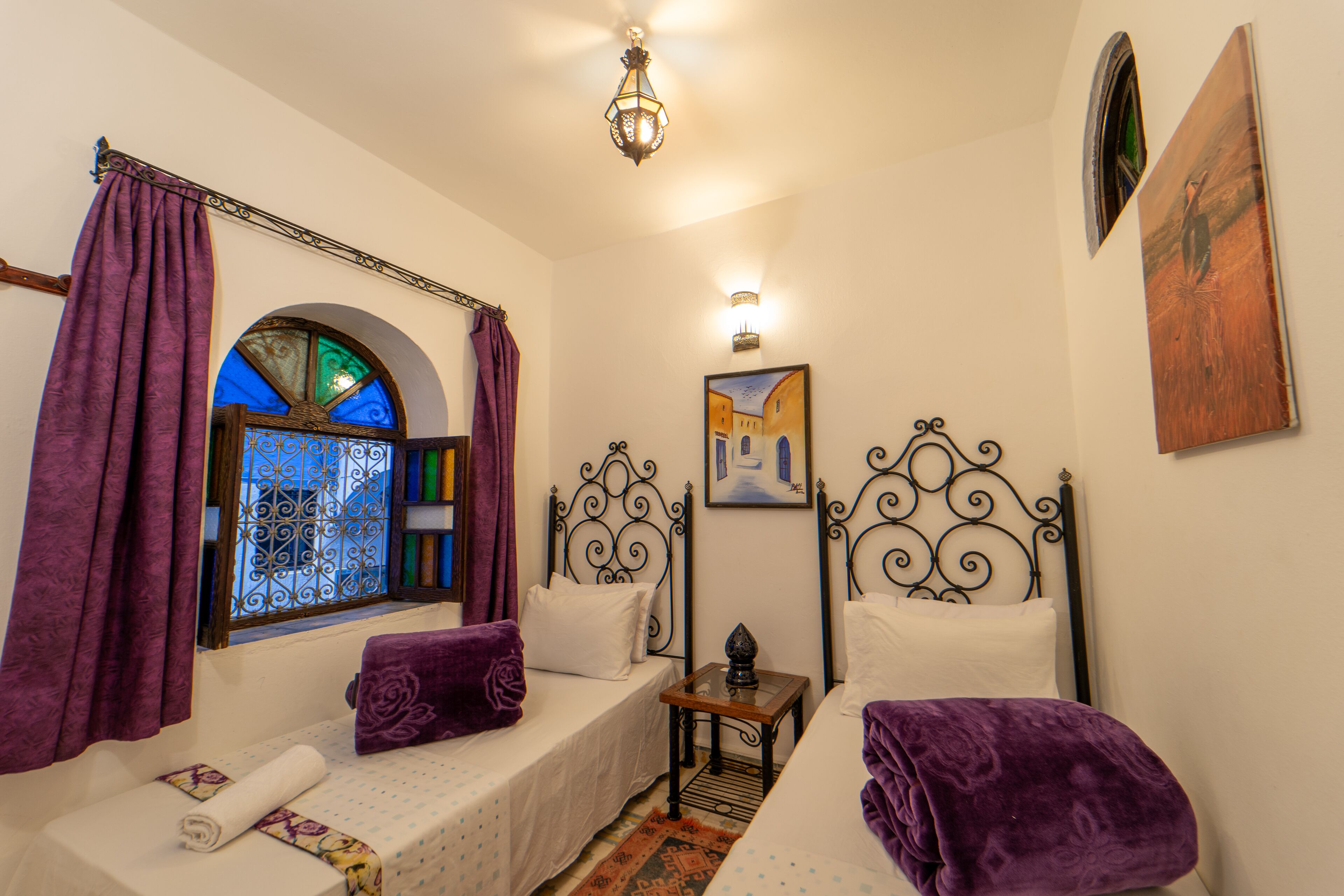 Quarto com duas camas, toalhas roxas, uma mesinha, um abajur decorativo, um quadro e um vitral.