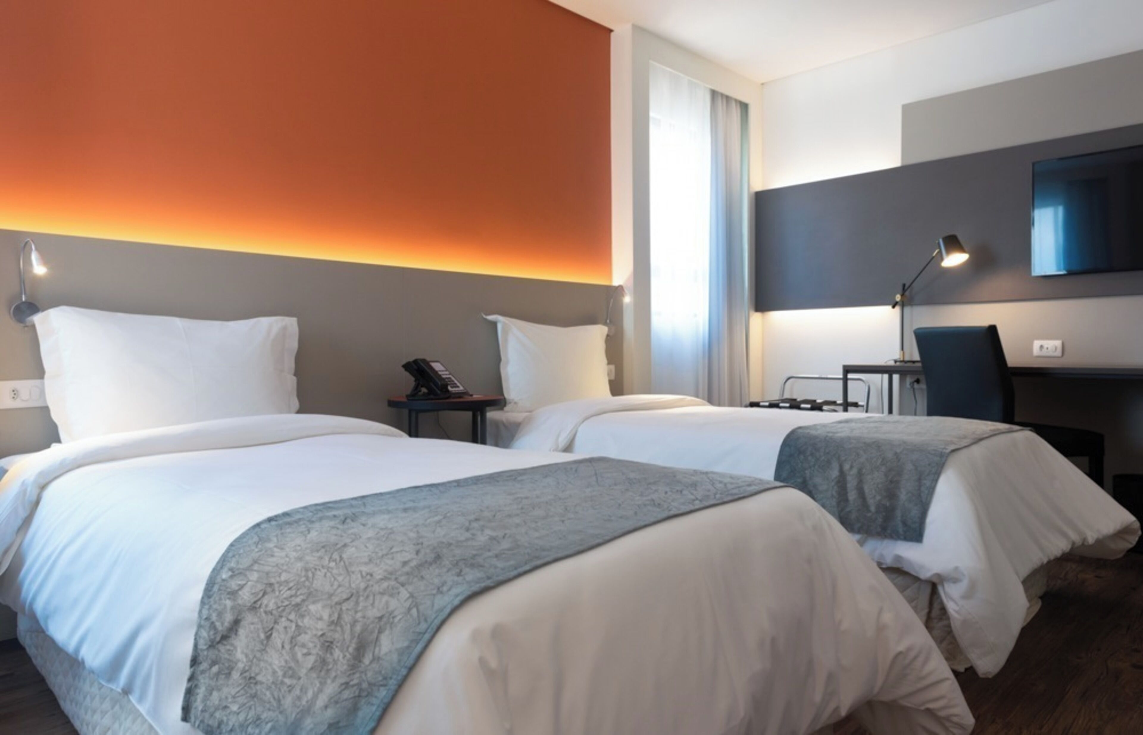 Quarto de hotel com duas camas, uma escrivaninha, uma cadeira, uma televisão e um telefone no criado-mudo.