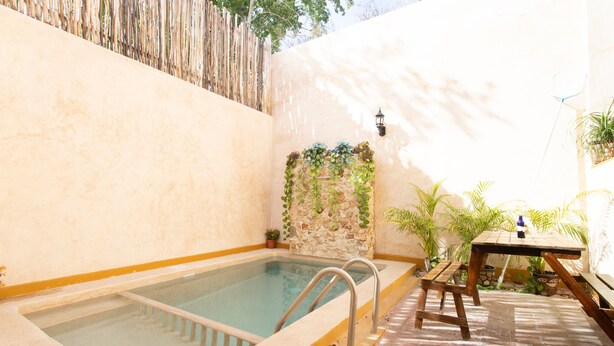 Piscina com parede de pedra e plantas, um banco de madeira e uma mesa com uma garrafa.