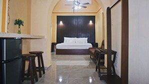 Quarto de hotel com cama, escrivaninha, dois banquetes, ventoinha no teto e uma janela.