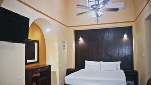 Quarto de hotel com uma cama grande, uma escrivaninha, uma cadeira, uma televisão e um ventilador de teto.