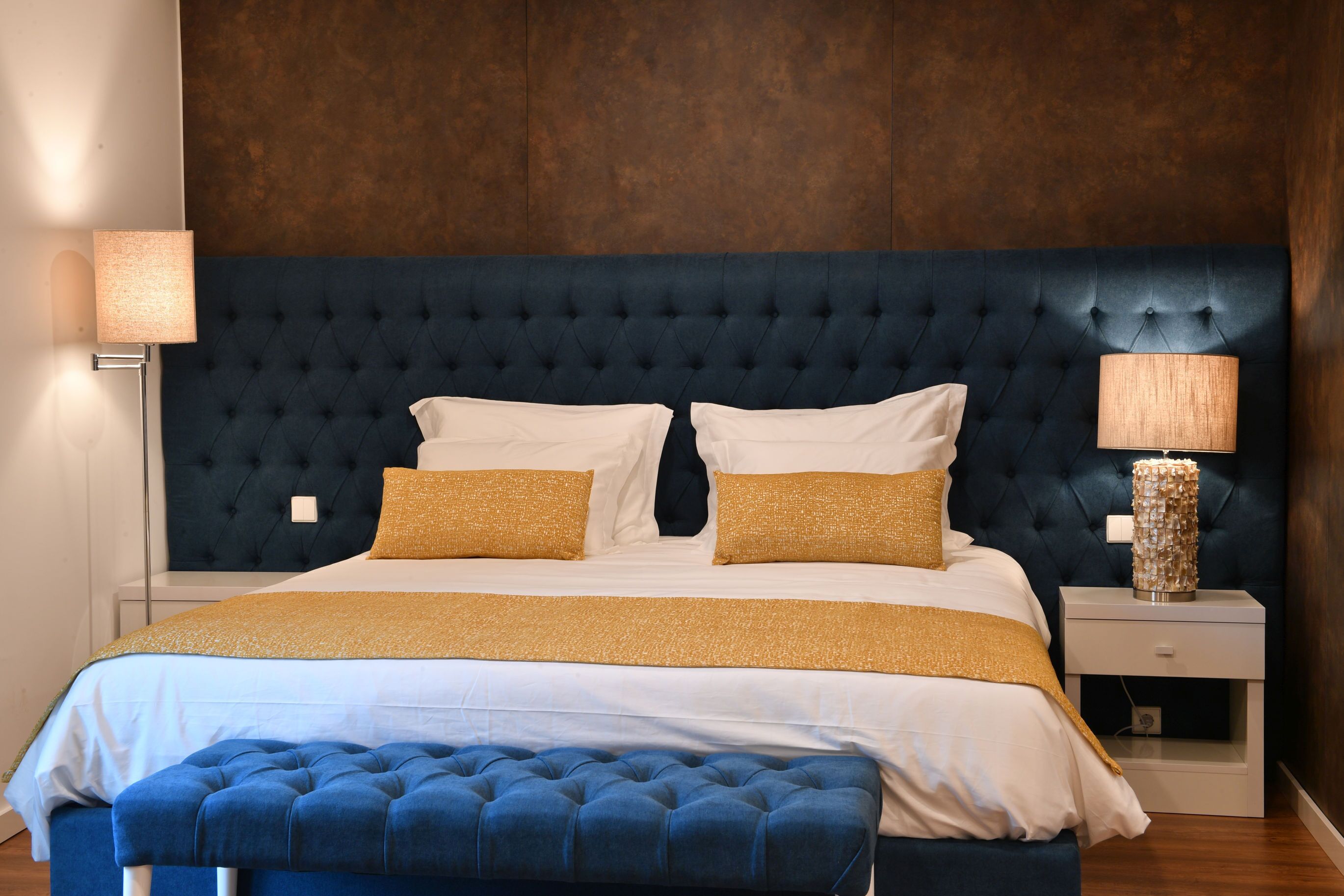 Quarto com cabeceira estofada, cama com roupa de cama branca e dourada, duas luminárias de mesa e um banquinho combinando.
