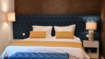 Quarto com cabeceira estofada, cama com roupa de cama branca e dourada, duas luminárias de mesa e um banquinho combinando.