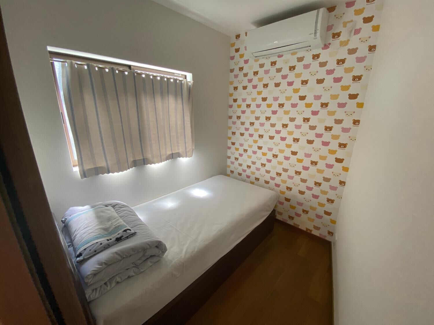 Quarto pequeno com cama de solteiro, janela, ventoinha no teto e parede com padrão de urso.