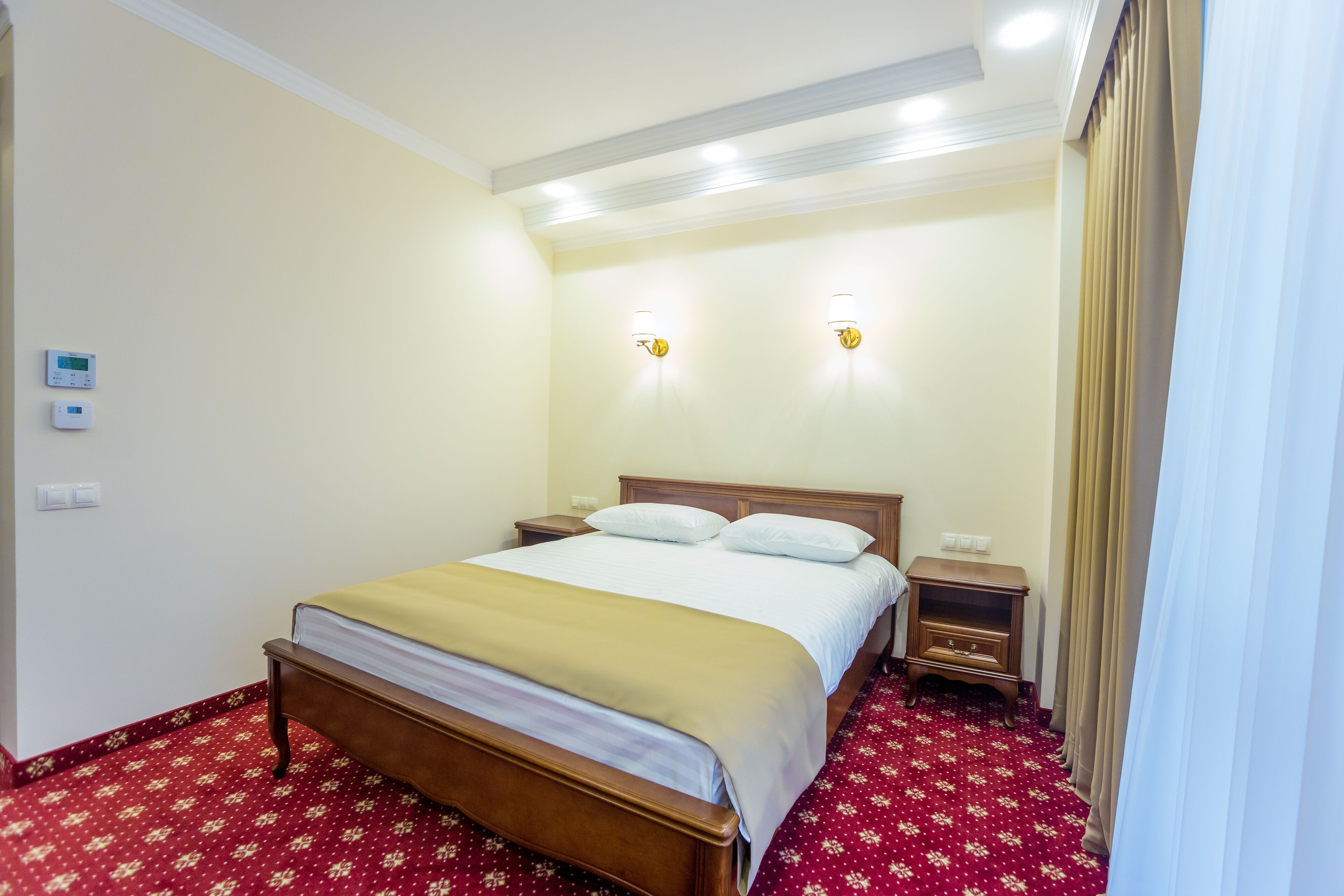 Quarto de hotel com cama, mesas de cabeceira, termostato fixado na parede e luminária de parede.