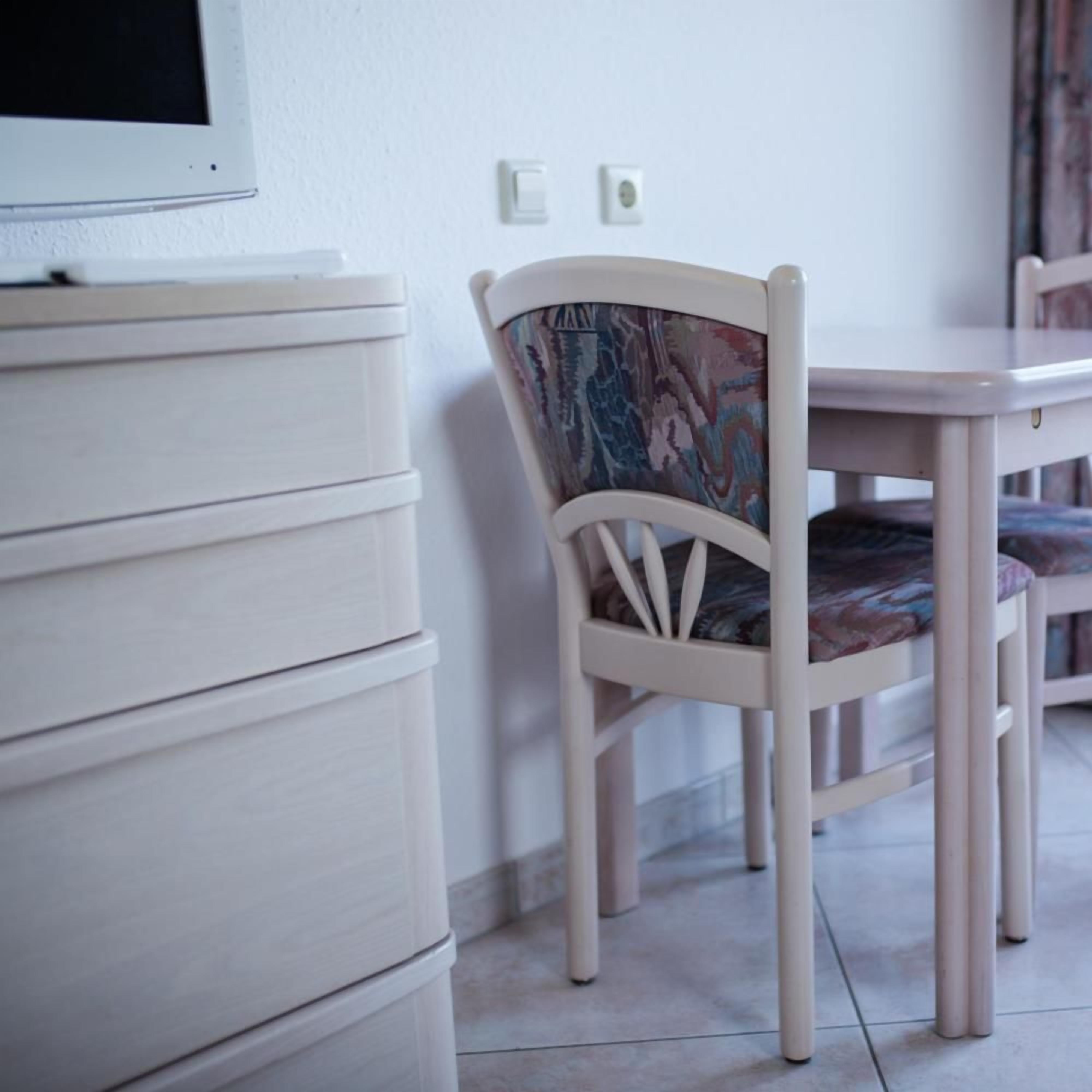 Quarto com cômoda branca, cadeira com almofada estampada e televisão na parede.