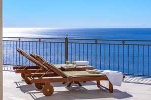 Uma varanda com duas poltronas de madeira, uma mesa com um copo de bebida e um livro, e vista para o mar.