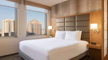 Quarto de hotel com uma cama grande, duas luminárias de cabeceira, uma mesinha de noite com relógio e vista para edifícios altos através de grandes janelas.