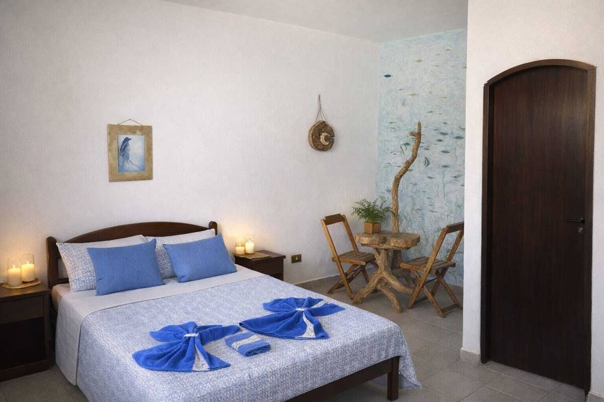 Quarto com cama, luminárias de cabeceira, uma mesinha com cadeiras, um relógio de parede e uma porta.