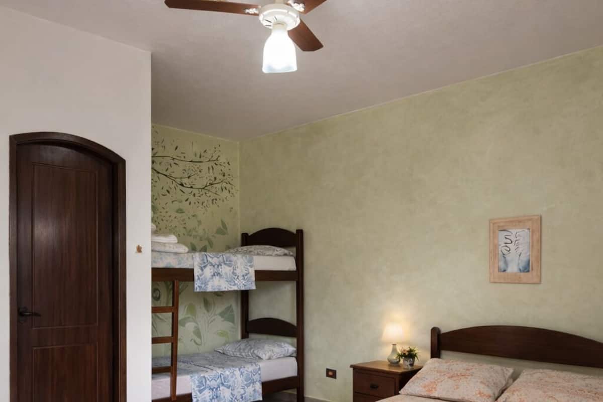 Quarto com cama, ventoinha no teto, mesa de cabeceira, guarda-roupa de madeira e beliche.