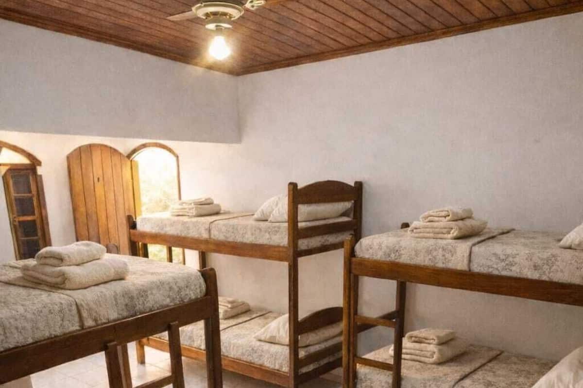 Quarto com quatro beliches de madeira, cada um com colchão e almofadas. Há um ventilador de teto e um luminário.