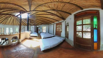 Quarto com telhado de palha, uma cama, uma mesinha e janela com vista para o verde.