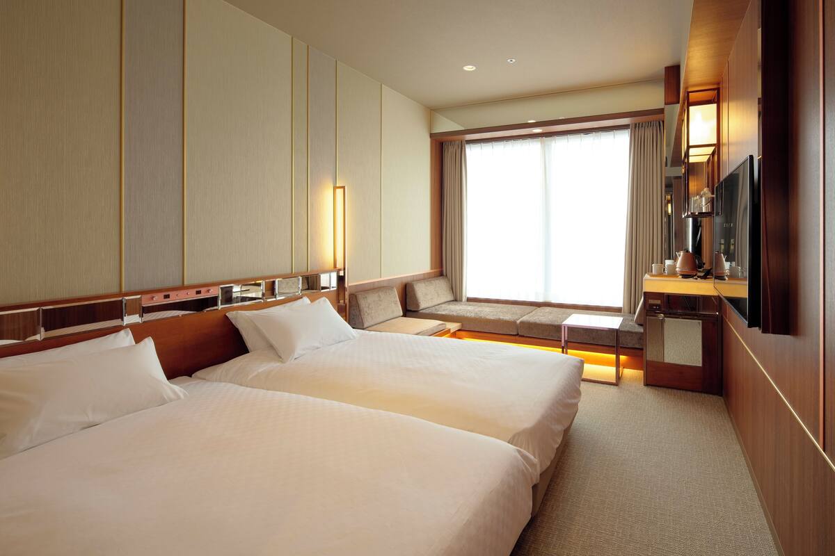 Quarto de hotel com duas camas, uma escrivaninha, televisão e uma janela grande com cortinas.