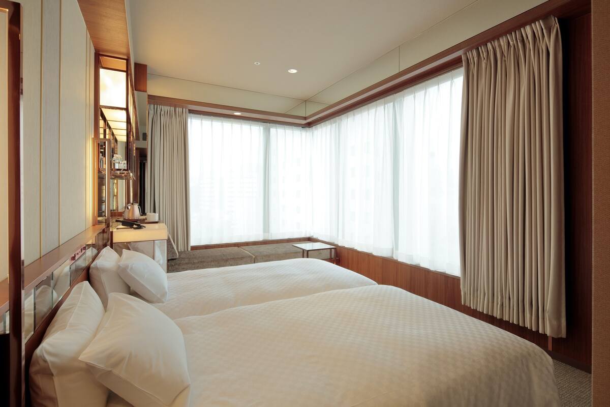 Quarto de hotel com duas camas, uma escrivaninha com luminária e janelas amplas com cortinas.