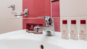 Uma casa de banho com uma pia branca, torneira cromada e paredes com azulejos vermelhos. Há três frascos de shampoo do hotel Madrid sobre a pia.