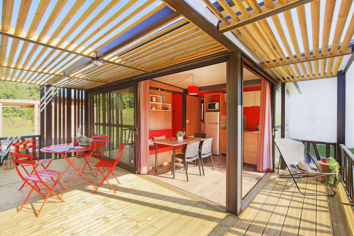 Uma sala de estar integrada com deck de madeira, uma mesa de jantar com cadeiras vermelhas e uma área de cozinha com eletrodoméstico vermelho.