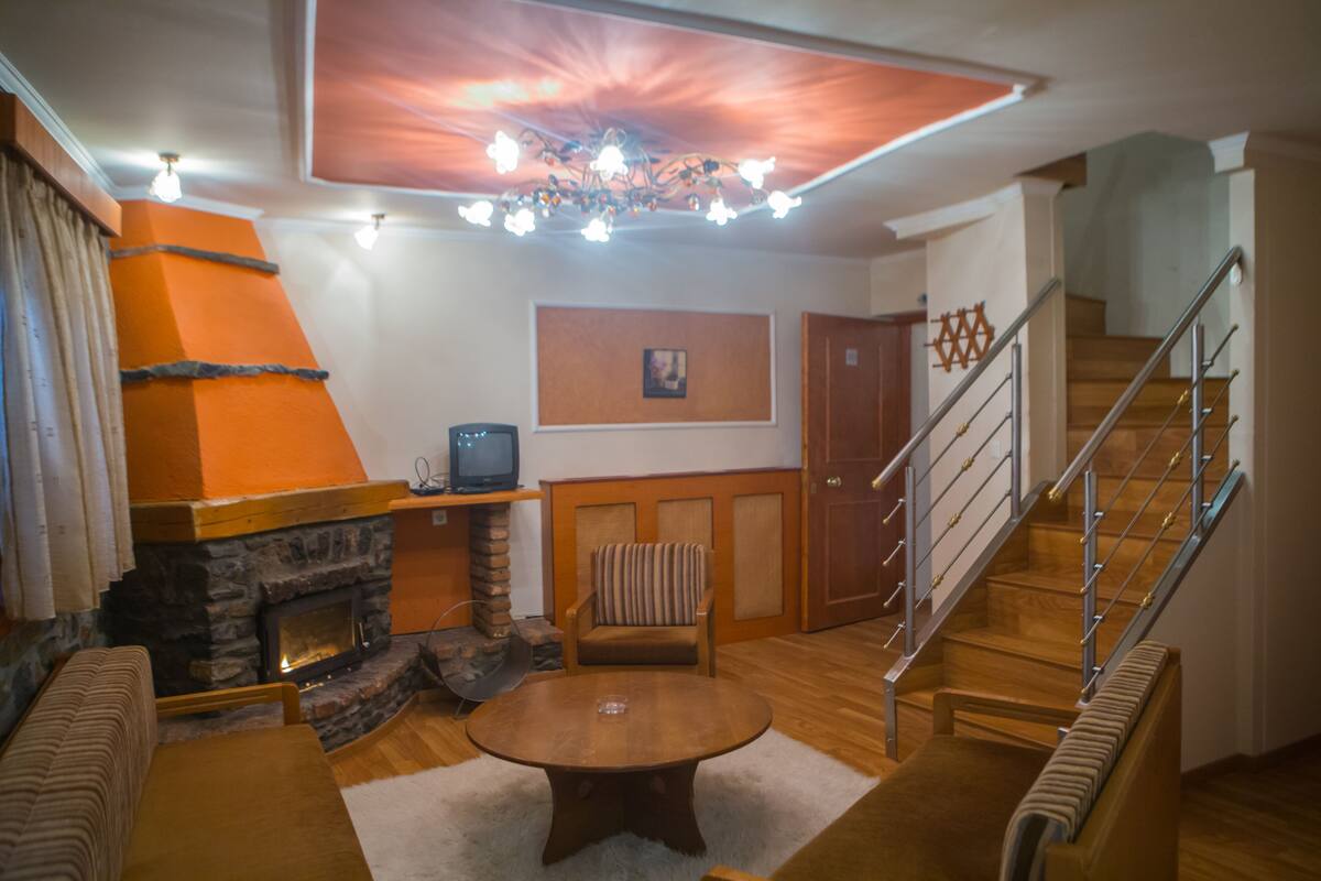Uma sala de estar com lareira de pedra, mesa de centro de madeira, poltrona listrada e escada com corrimão de metal.