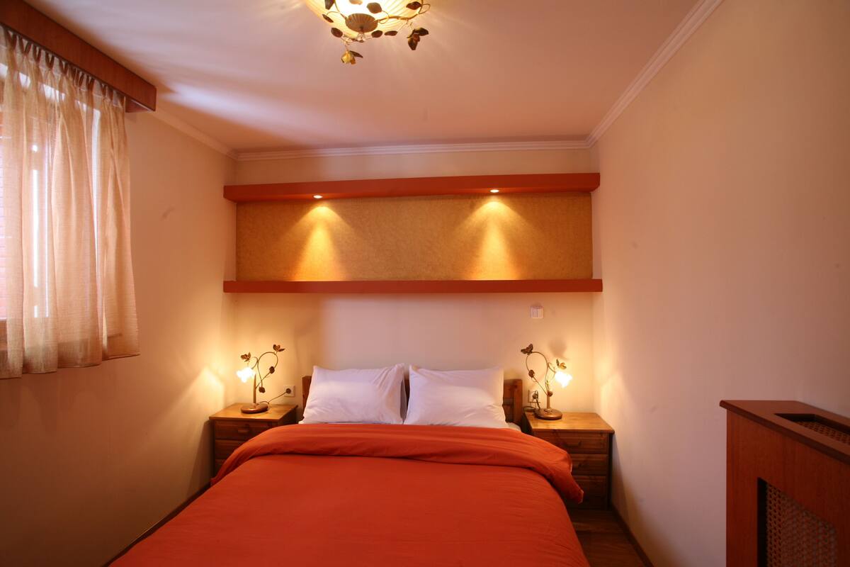 Quarto com uma cama, duas mesas de cabeceira com luminárias, um candeeiro no teto e uma janela com cortinas.