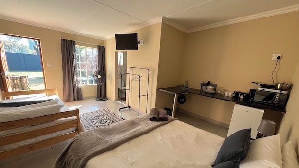 Quarto de hotel com cama, pequeno espaço de cozinha, televisão e janela com vista para árvores.