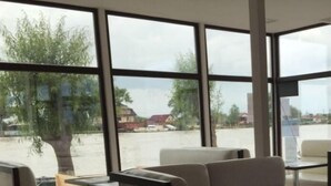 Interior moderno com grandes janelas, área de estar com sofás e uma mesa, e vista para a água e edifícios.