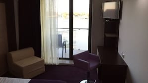 Quarto de hotel com cama, sofá, poltrona, televisão e porta de vidro deslizante para o exterior.