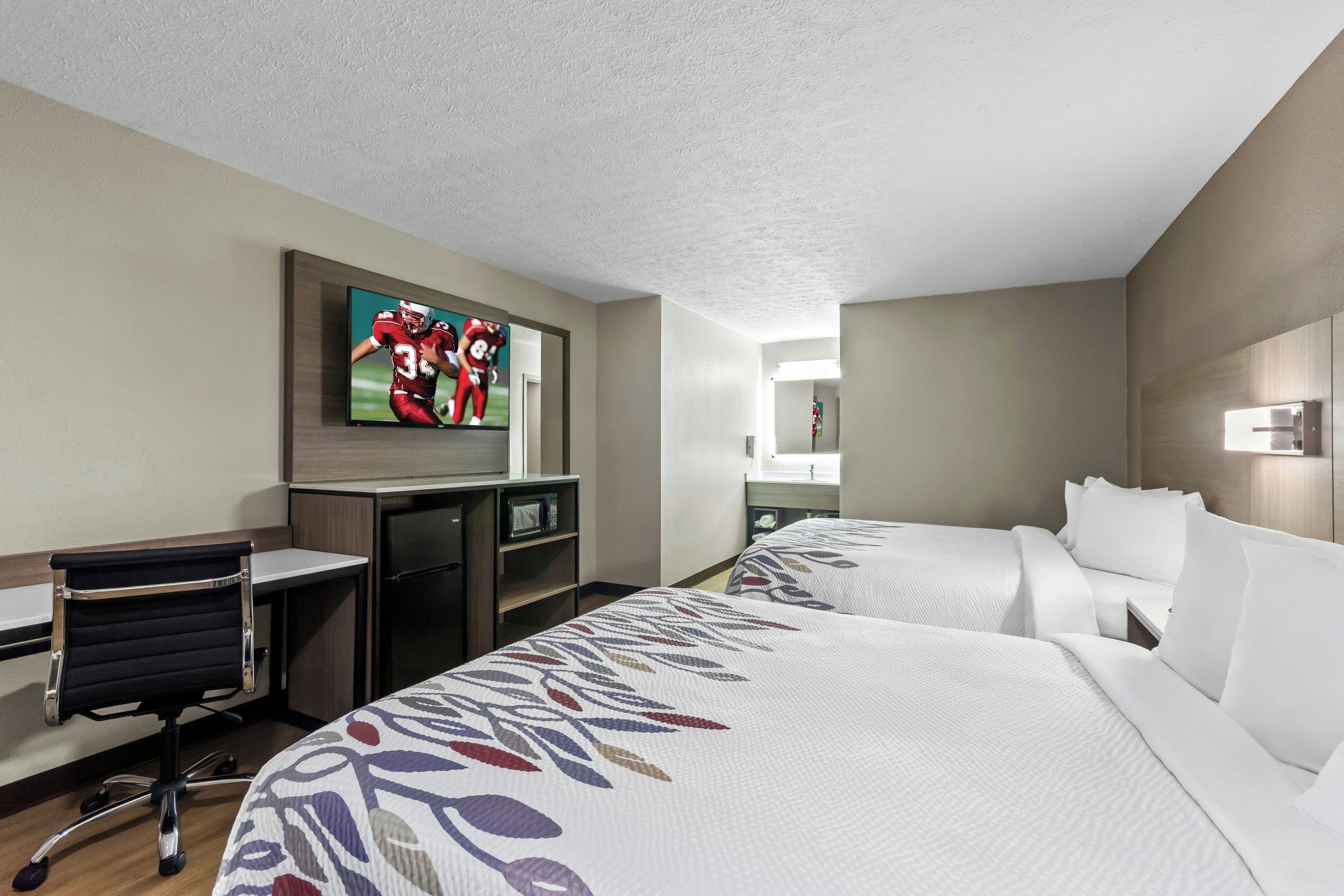 Quarto de hotel com uma cama, uma escrivaninha, uma cadeira, uma televisão e um espelho.