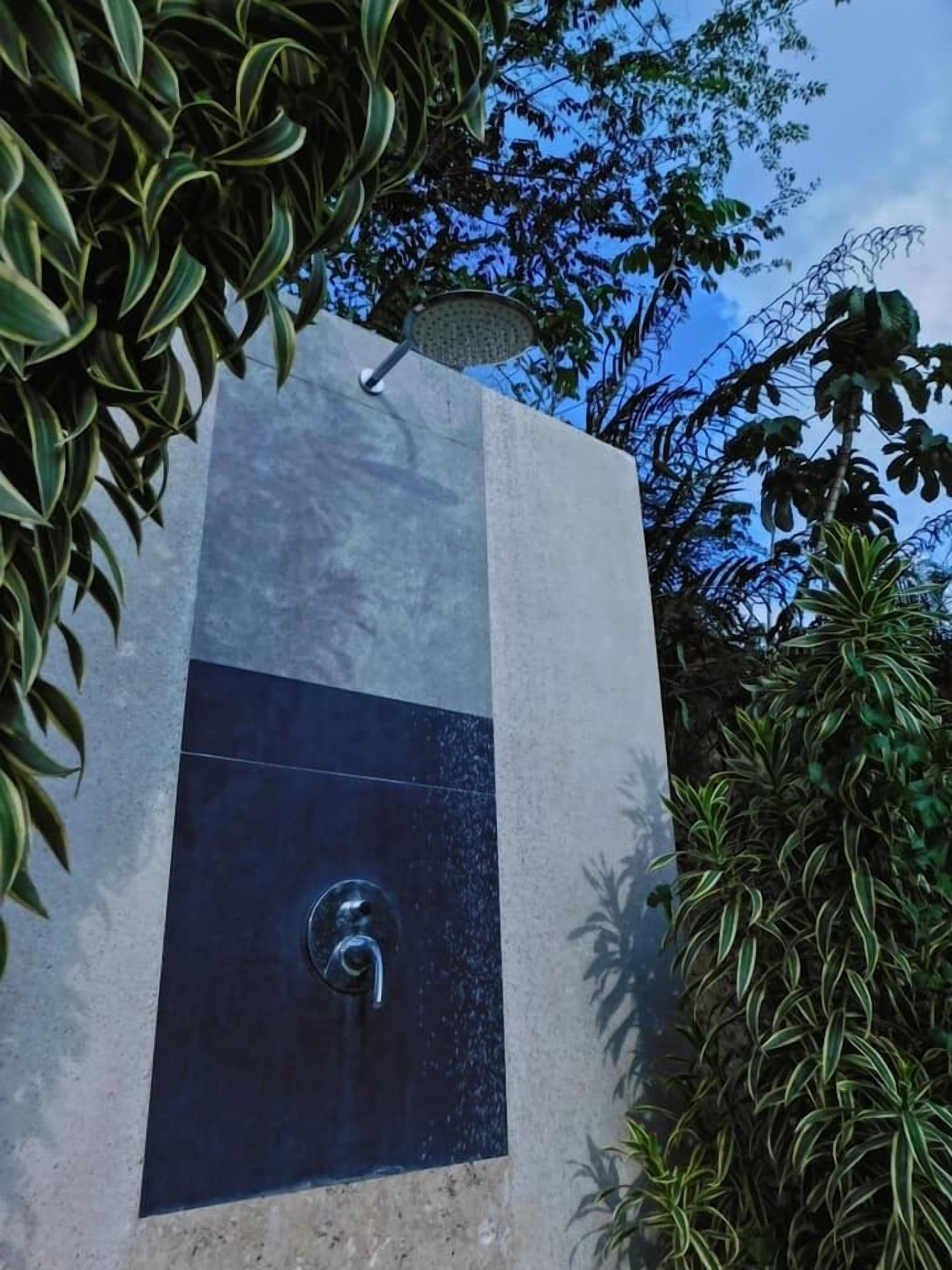 Uma fonte moderna ao ar livre com um bico de metal, rodeada por vegetação exuberante e um céu azul límpido.