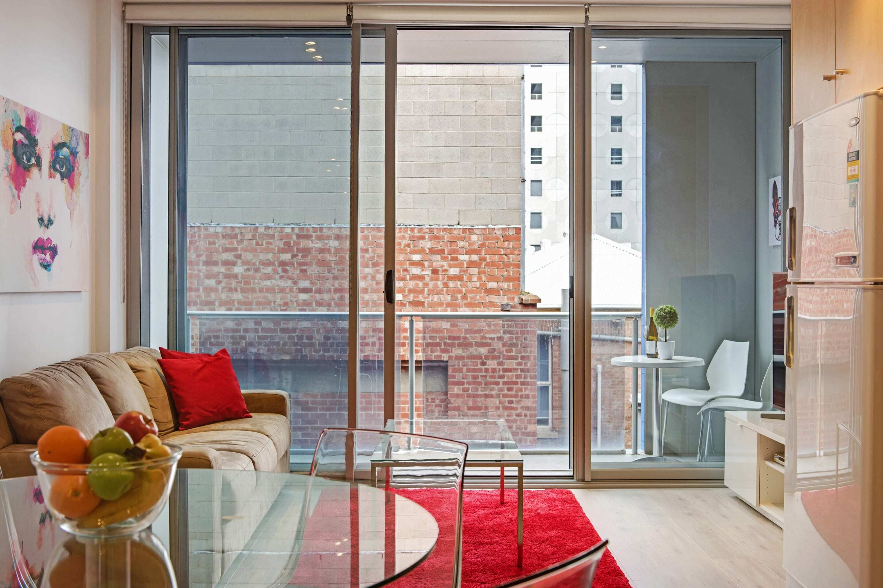 Uma sala de estar moderna com uma mesa de vidro, um tapete vermelho, um sofá com almofada vermelha e uma janela grande com vista para um edifício de tijolos.