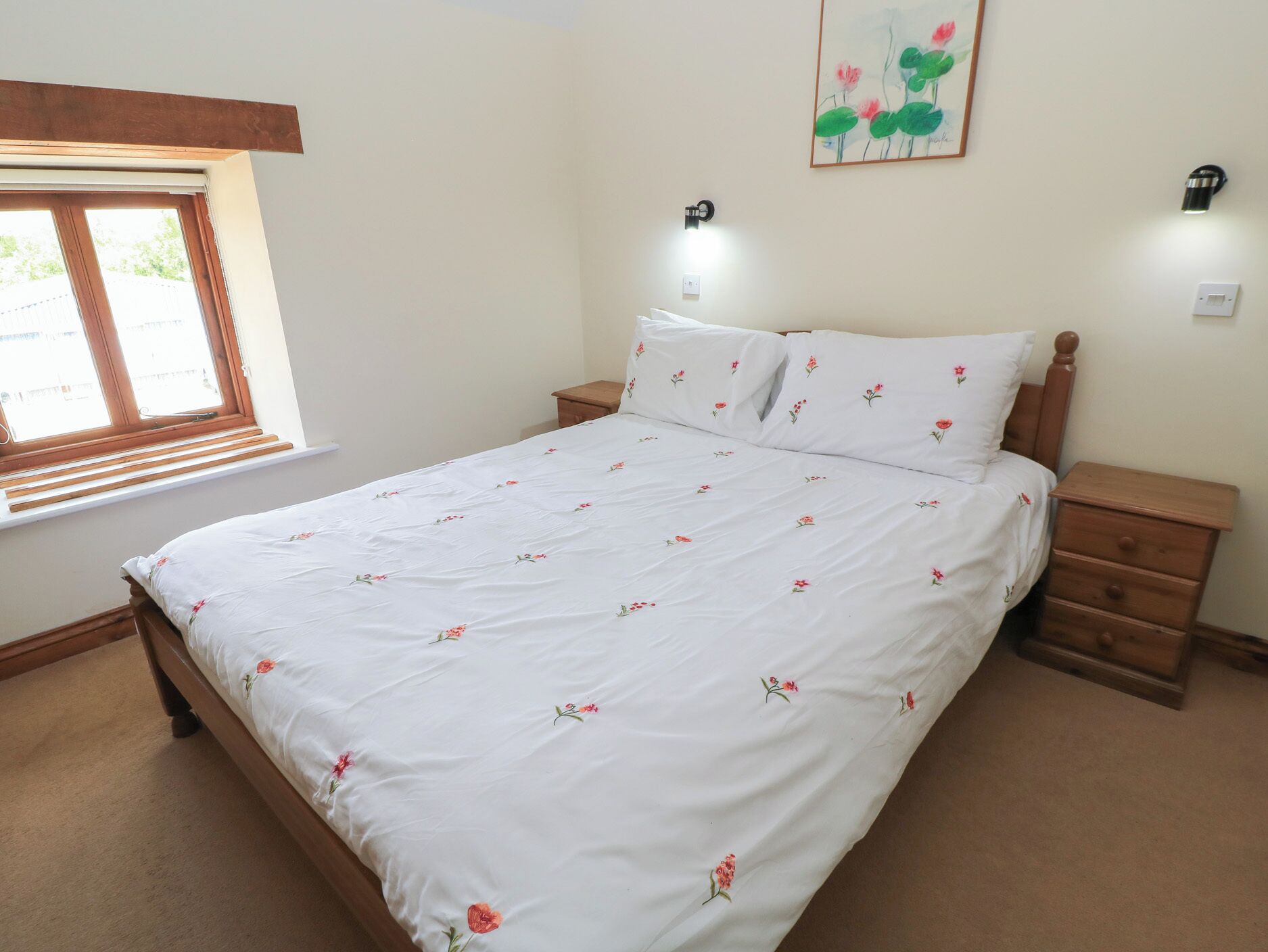 Um quarto com cama, cabeceira de madeira, duas mesas de cabeceira, um criado-mudo, janela com persianas e luminária fixada na parede.