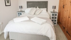 Cama bem arrumada com lençóis brancos e duas toalhas, cabeceira cinza e duas mesas de cabeceira com abajures.
