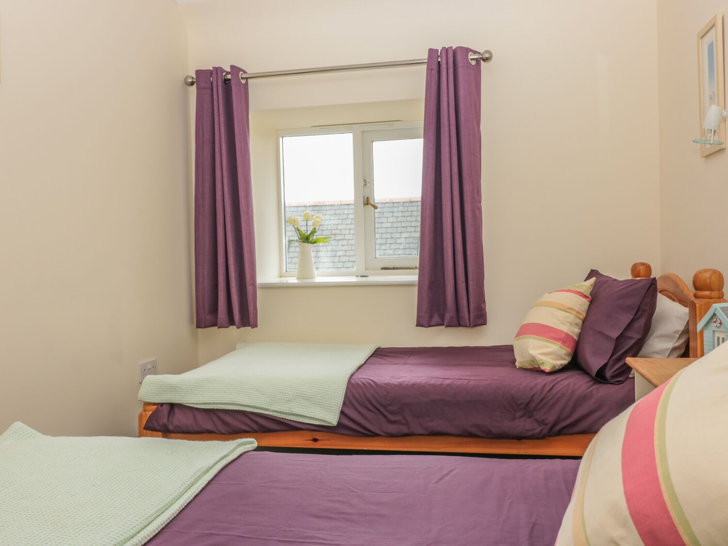 Quarto com duas camas, roupa de cama roxa, janela com cortinas, mesa de cabeceira com abajur e uma pequena planta no parapeito.