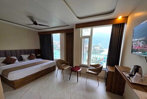 Quarto de hotel com cama, duas cadeiras, uma mesinha, escrivaninha com cafeteira e uma janela grande com vista para a cidade e montanhas.