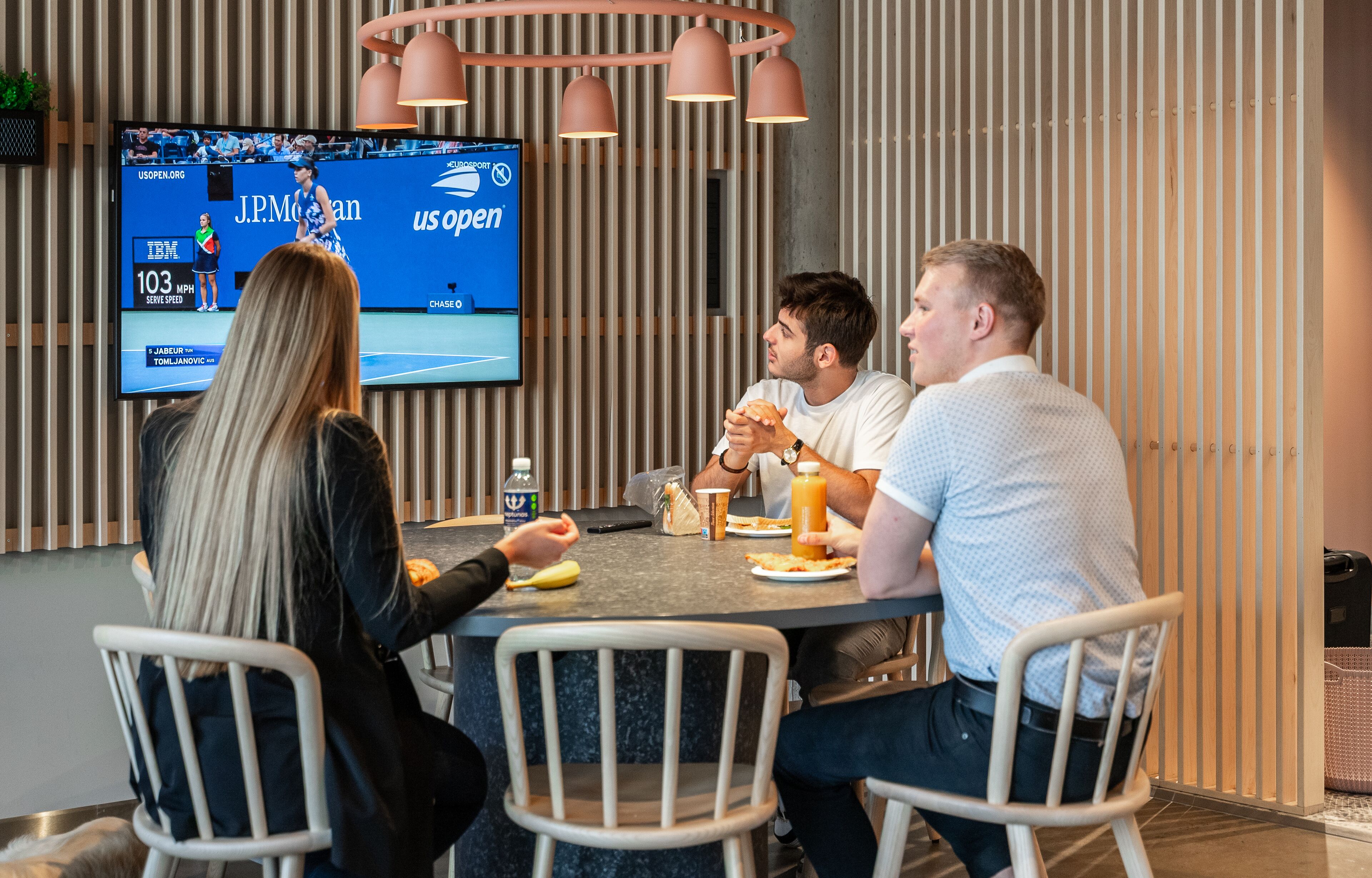 Um grupo de pessoas jantando em um restaurante moderno com uma grande tela exibindo uma partida de tênis.