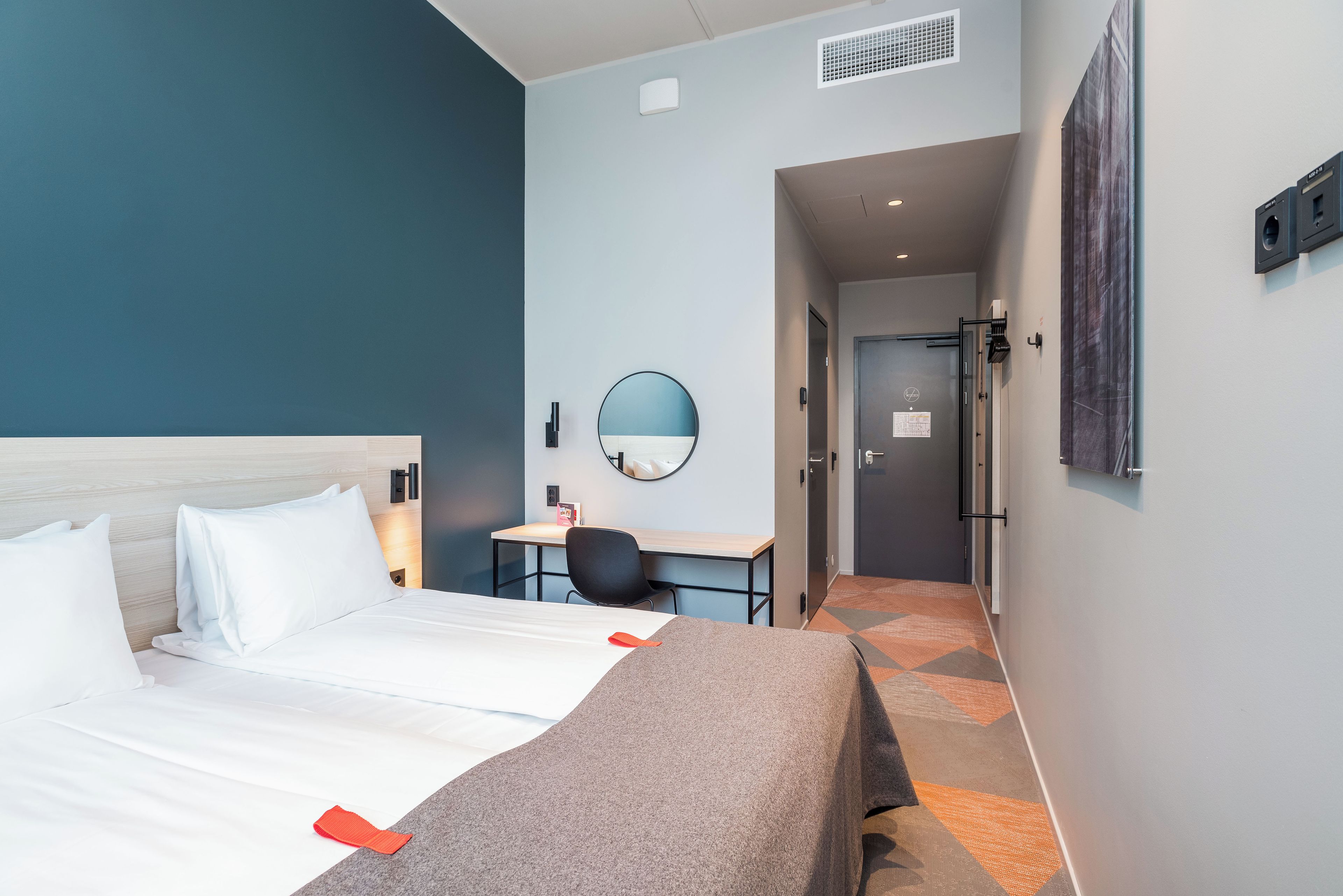 Quarto de hotel com cama, escrivaninha, cadeira, espelho e porta que dá para outro cômodo.