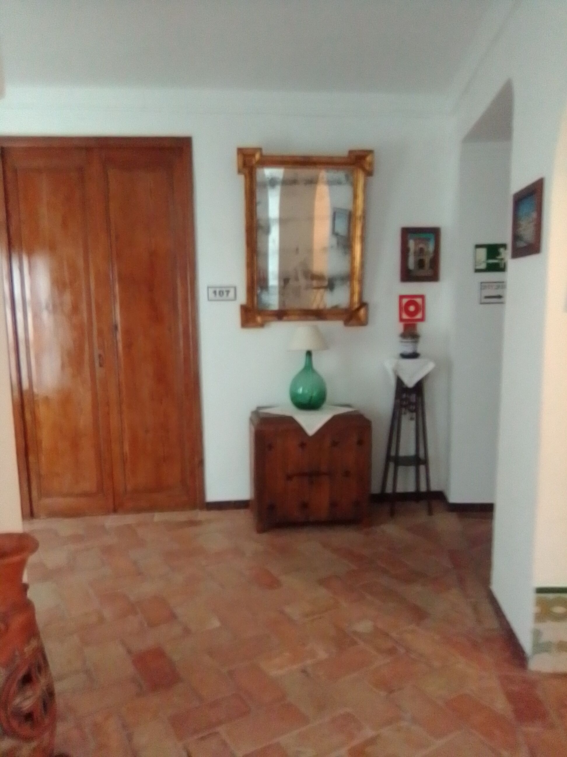 Quarto com porta de madeira, espelho, mesa pequena com luminária e chão de azulejos.