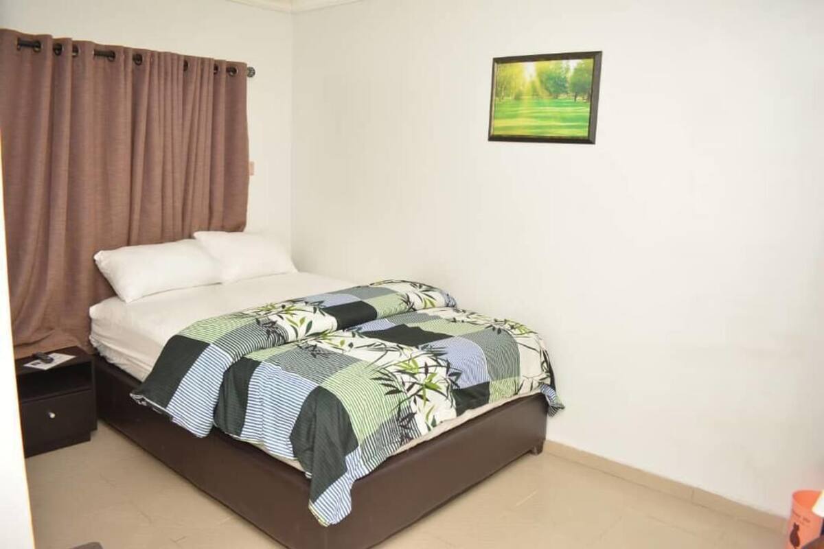 Quarto com cama, cortinas castanhas, mesa de cabeceira, quadro na parede e chão de azulejos.