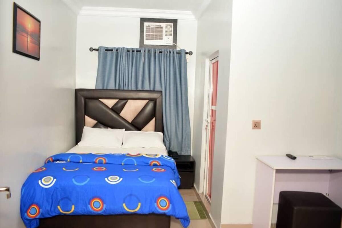 Um quarto pequeno e bem organizado, com uma cama, uma escrivaninha e uma cadeira.