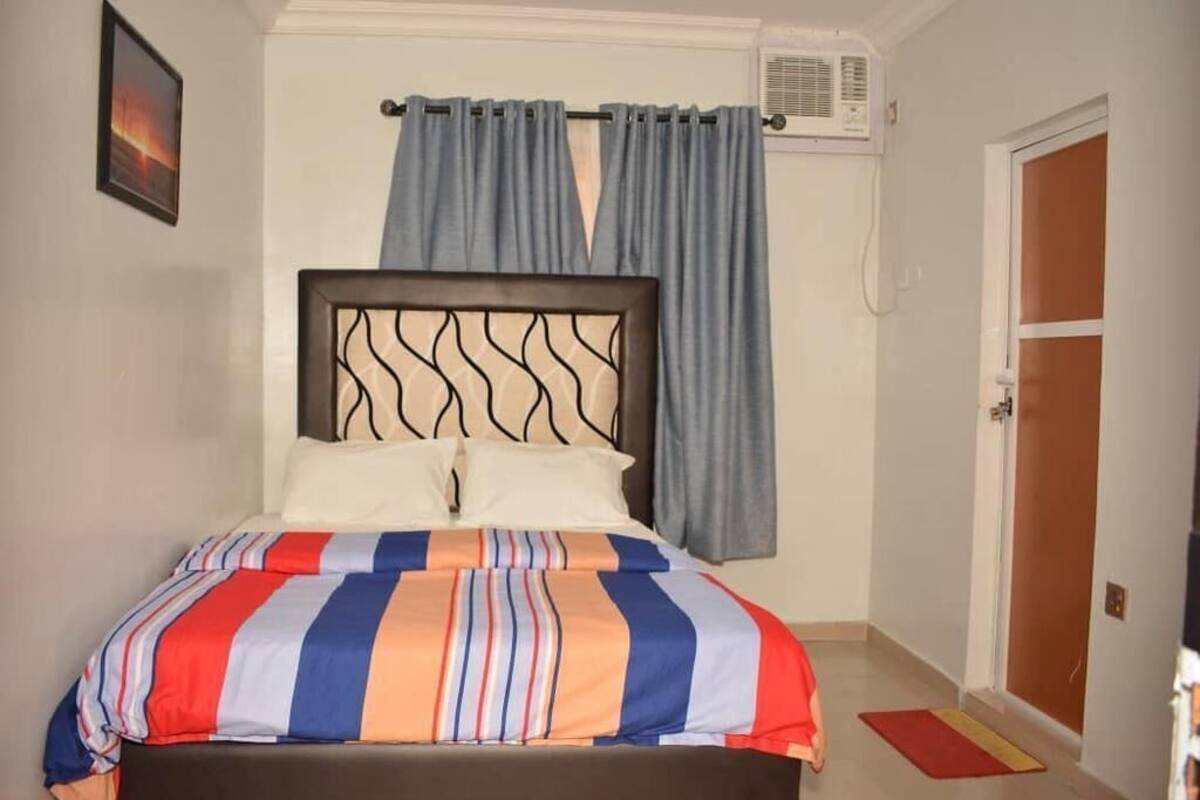 Uma cama com colcha listrada, cabeceira com design ondulado, cortina cinza e porta com tapete vermelho.