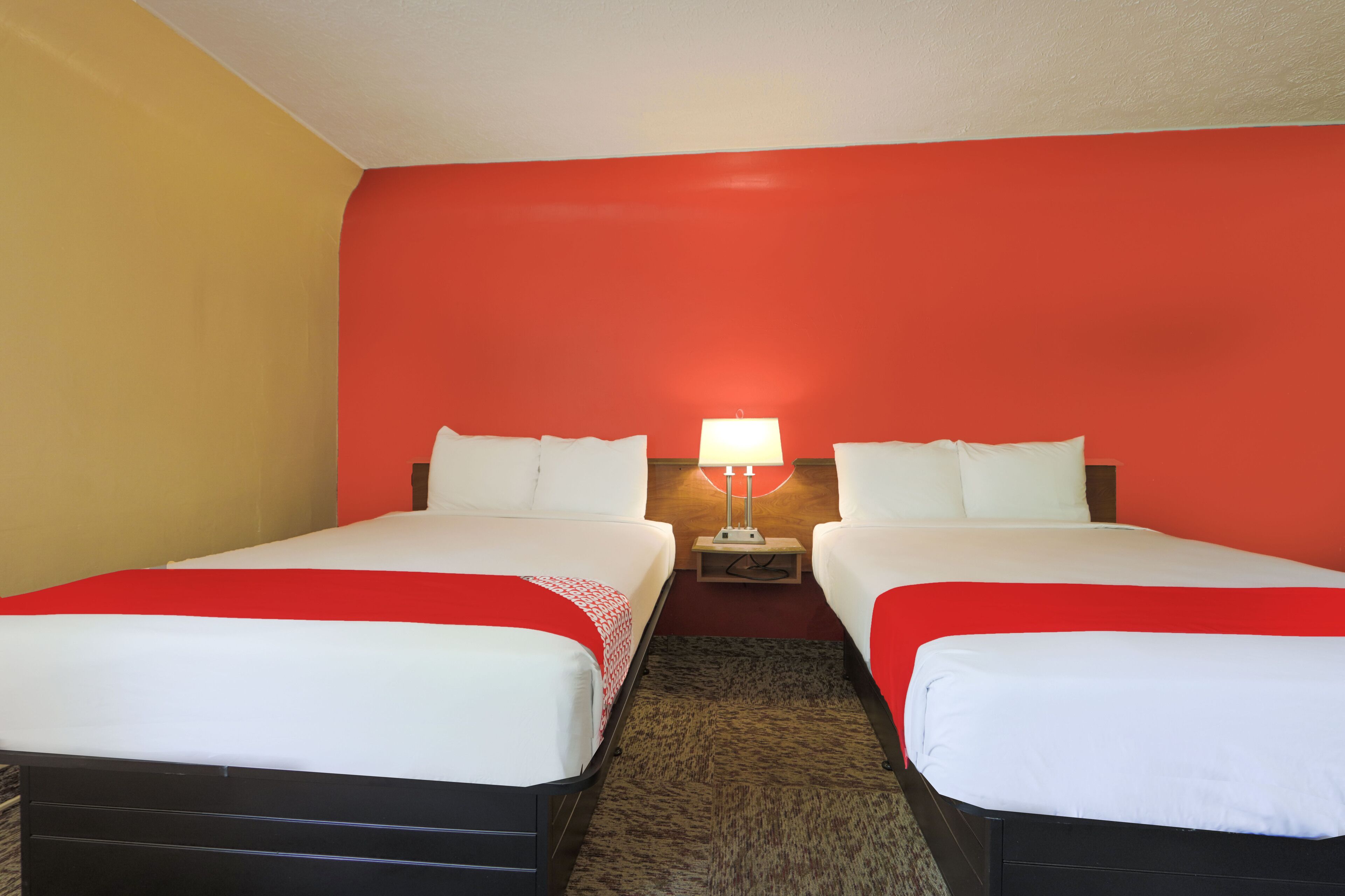 Quarto de hotel com duas camas, parede de destaque vermelha, mesa de cabeceira pequena com abajur e piso coberto por carpete.