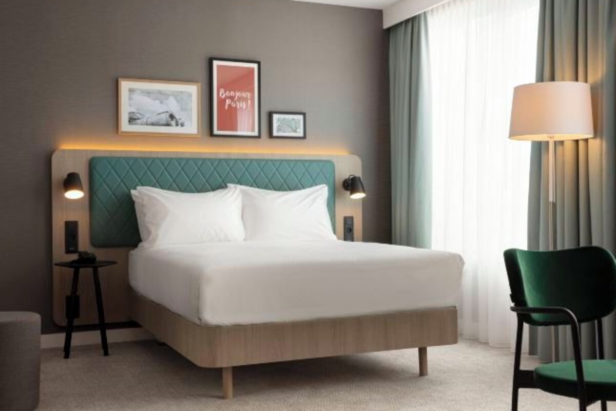 Quarto de hotel com uma cama, duas mesas de cabeceira, uma cadeira verde, uma luminária e quadros nas paredes.
